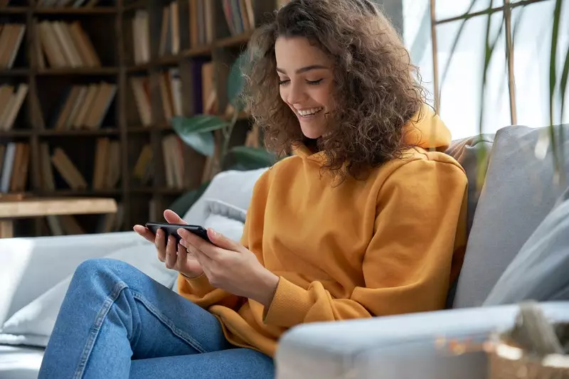 Een jonge vrouw in een geel sweatshirt kijkt naar haar telefoon en glimlacht.