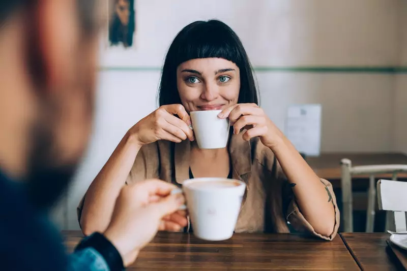 Wong wadon sing seneng mesem lan nyekel cangkir kopi nalika ndeleng pasangane ing meja.