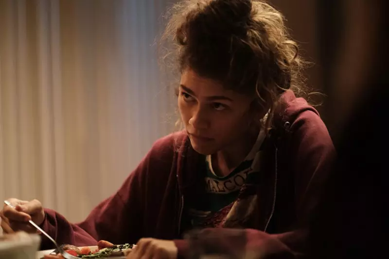 Zendaya als Rue in „Euphoria“. Sie trägt ihren charakteristischen roten Hoodie und sitzt am Küchentisch …